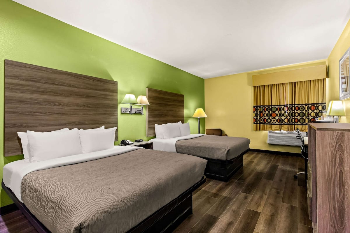 Quarto de hotel com duas camas, cabeceira de madeira, uma escrivaninha com abajur e uma janela com cortinas.