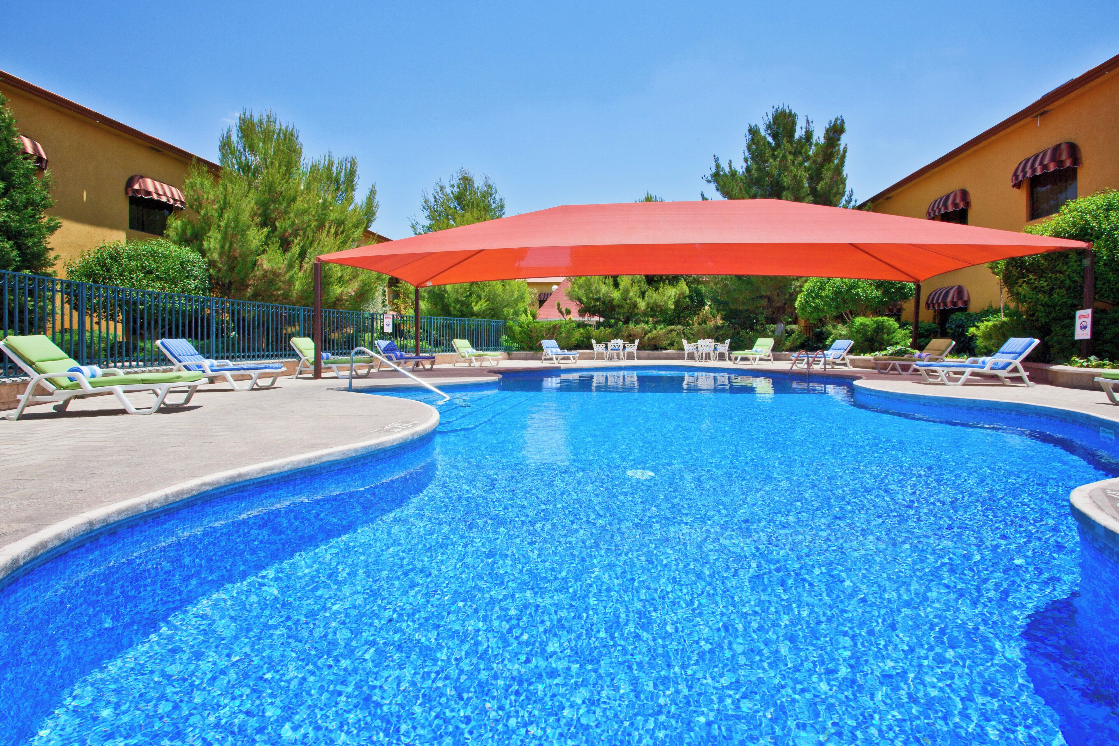 Piscina com várias espreguiçadeiras e guarda-sóis, cercada por áreas verdes e edifícios.
