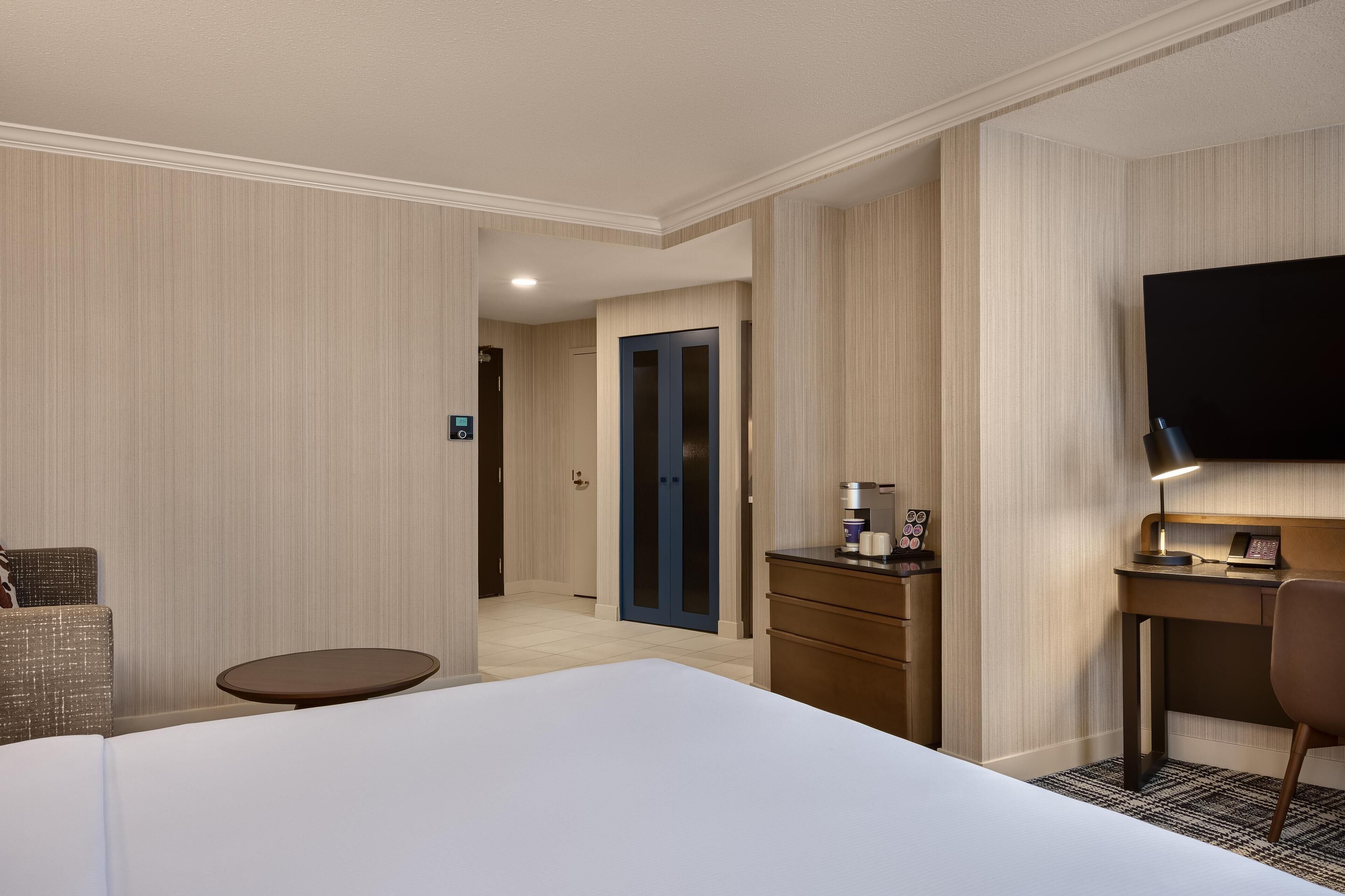 Quarto de hotel com cama, escrivaninha, cadeira, televisão e mesa pequena.