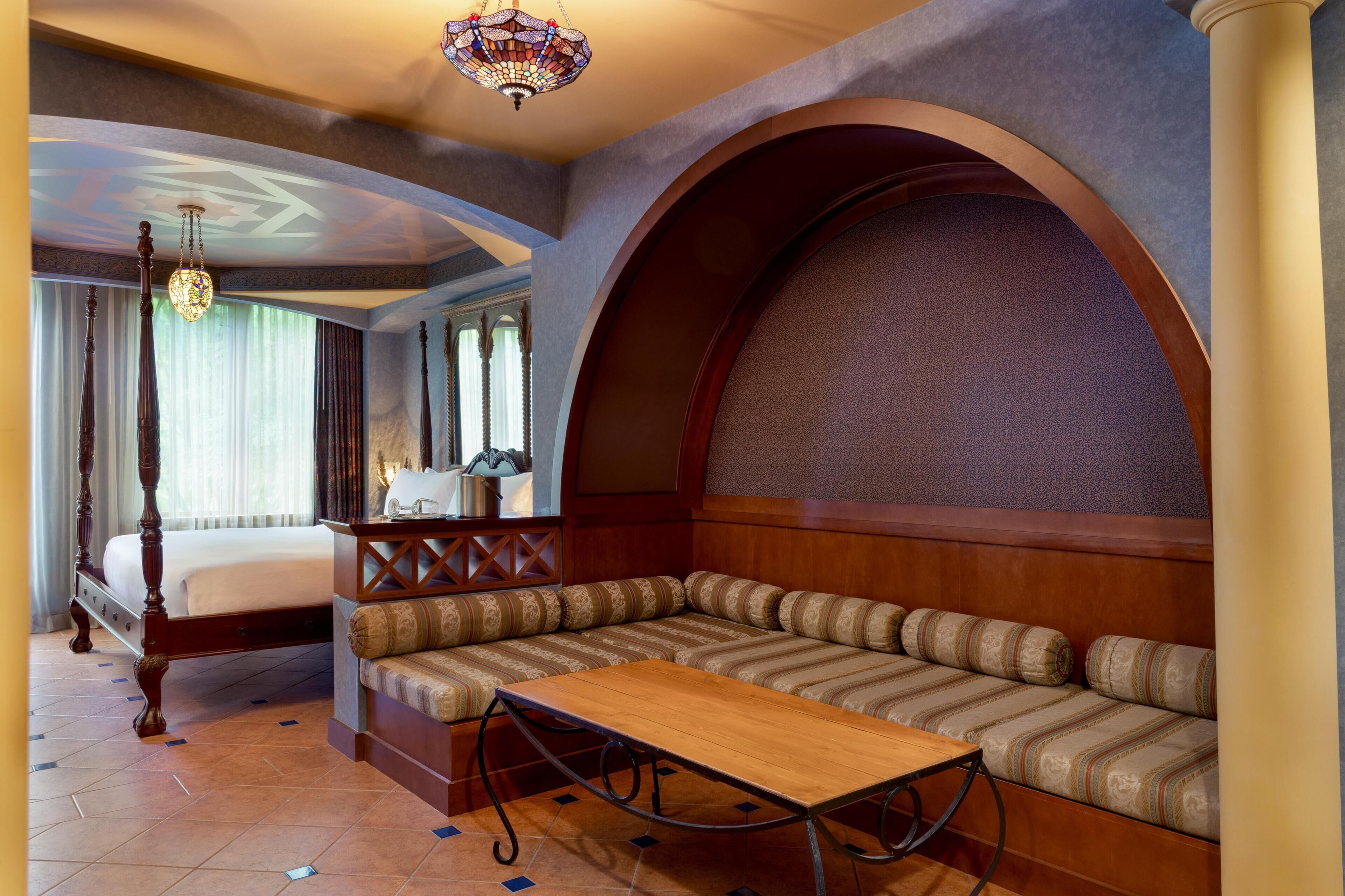 Quarto com cama de dossel, um banco com almofadas, uma mesa de madeira e uma luminária decorativa no teto.