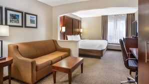 Quarto de hotel com sofá marrom, mesa de centro de madeira, cama com lençóis brancos, escrivaninha com cadeira, televisão e quadros nas paredes.