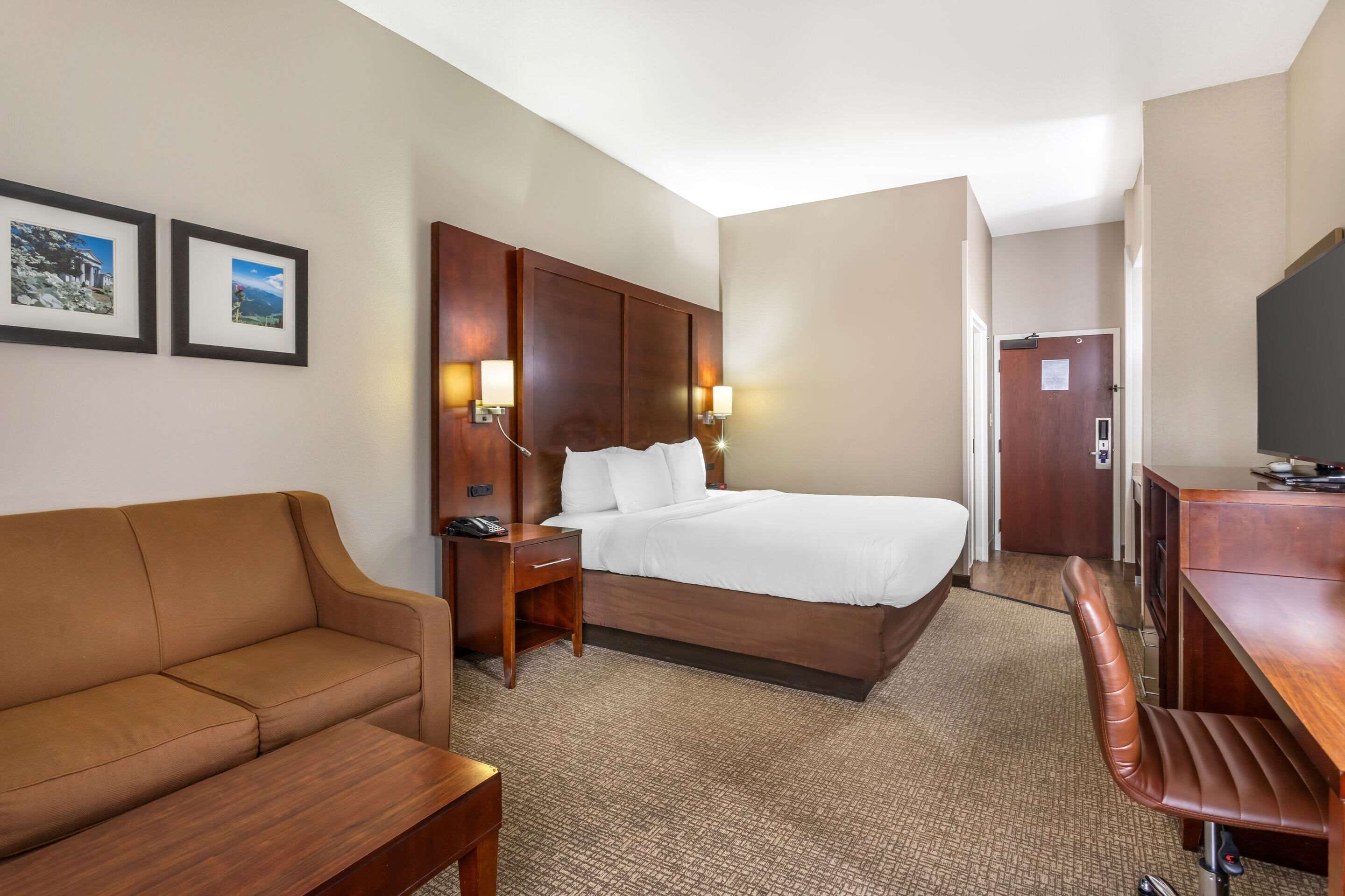 Quarto de hotel com cama, cabeceira de madeira, escrivaninha, poltrona de couro marrom, mesa de cabeceira de madeira e duas fotos emolduradas na parede.