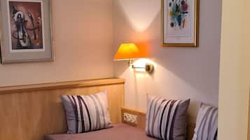 Quarto de hotel compacto com uma cama de solteiro, escrivaninha, cadeira e mesa de cabeceira.