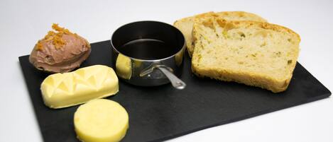 Uma bandeja de ardósia preta com três tipos de manteiga, um pequeno pote preto e duas fatias de pão.