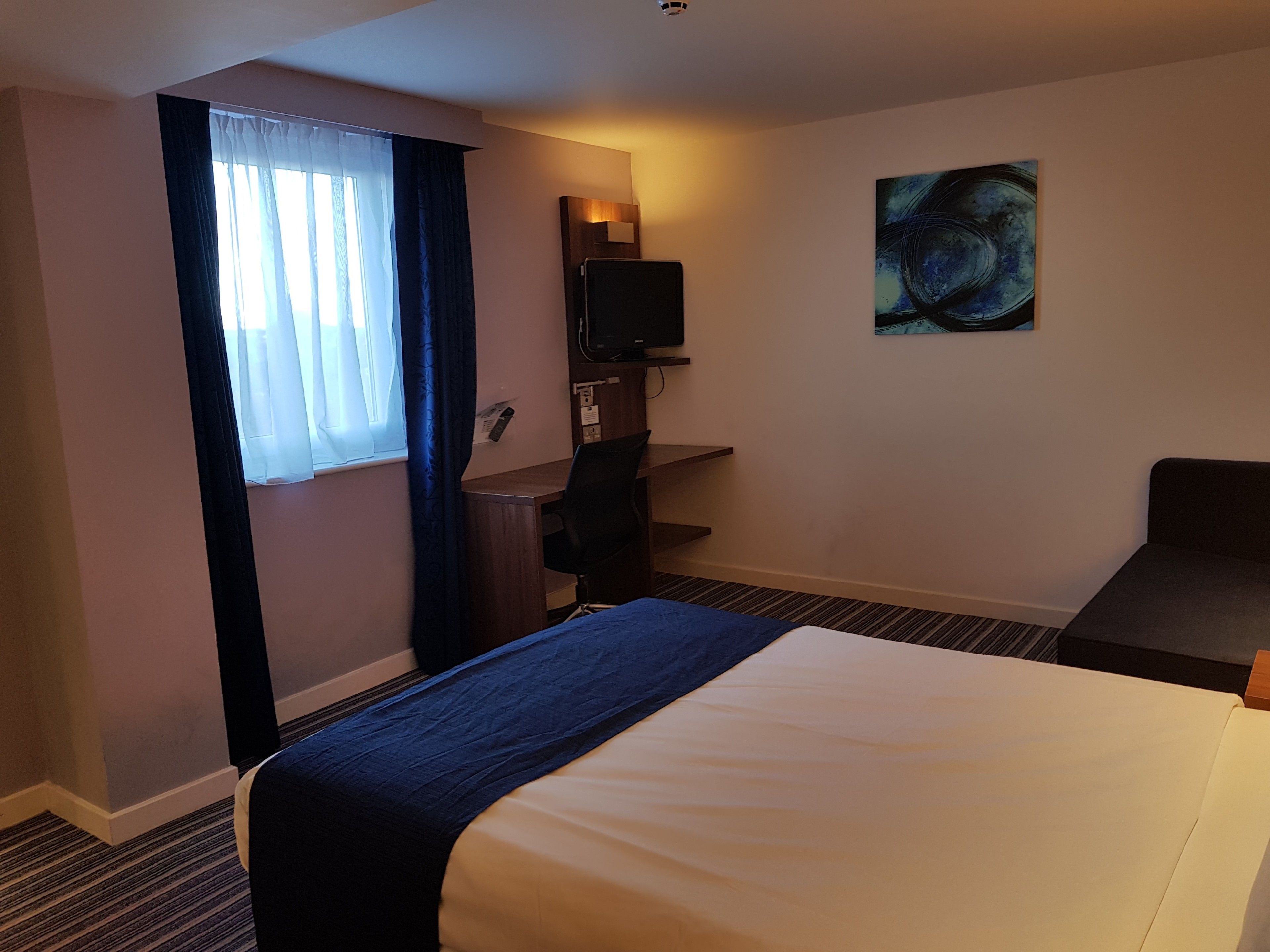 Quarto de hotel com cama, escrivaninha, cadeira, televisão e um quadro na parede.