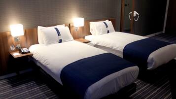 Quarto de hotel com duas camas, cada uma com edredão branco e almofadas azuis, mesa de cabeceira com telefone e luminária, e luz instalada na parede.