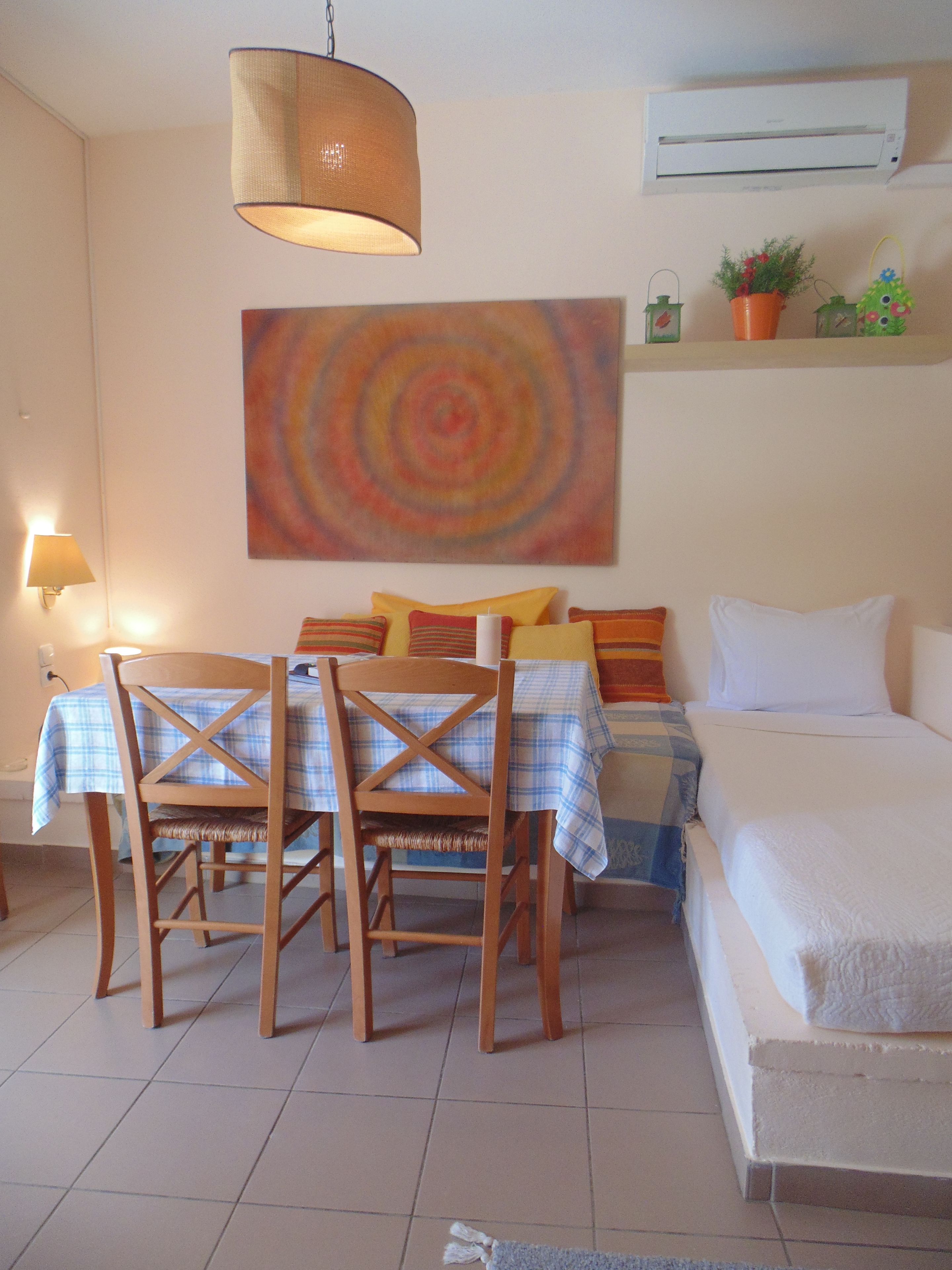 Quarto com mesa de jantar, cadeiras, cama, ar-condicionado fixado na parede e um quadro abstrato colorido.