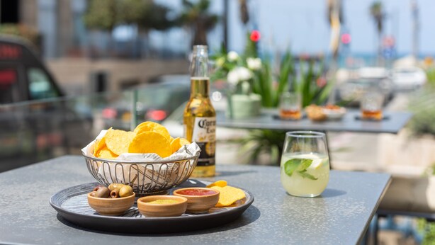 Uma mesa com petiscos, uma garrafa de cerveja e uma bebida num terraço com vista para o mar e edifícios.