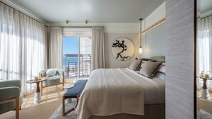 Quarto de hotel moderno com uma cama grande, área de estar com um banco e cadeiras, vista para o mar e um painel decorativo na parede.