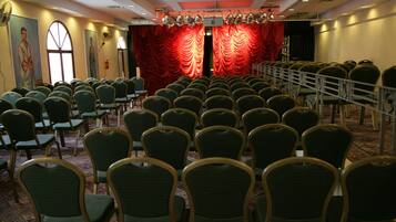 Uma sala de conferências com fileiras de cadeiras, um palco com cortinas vermelhas e um grande ventilador na parede.
