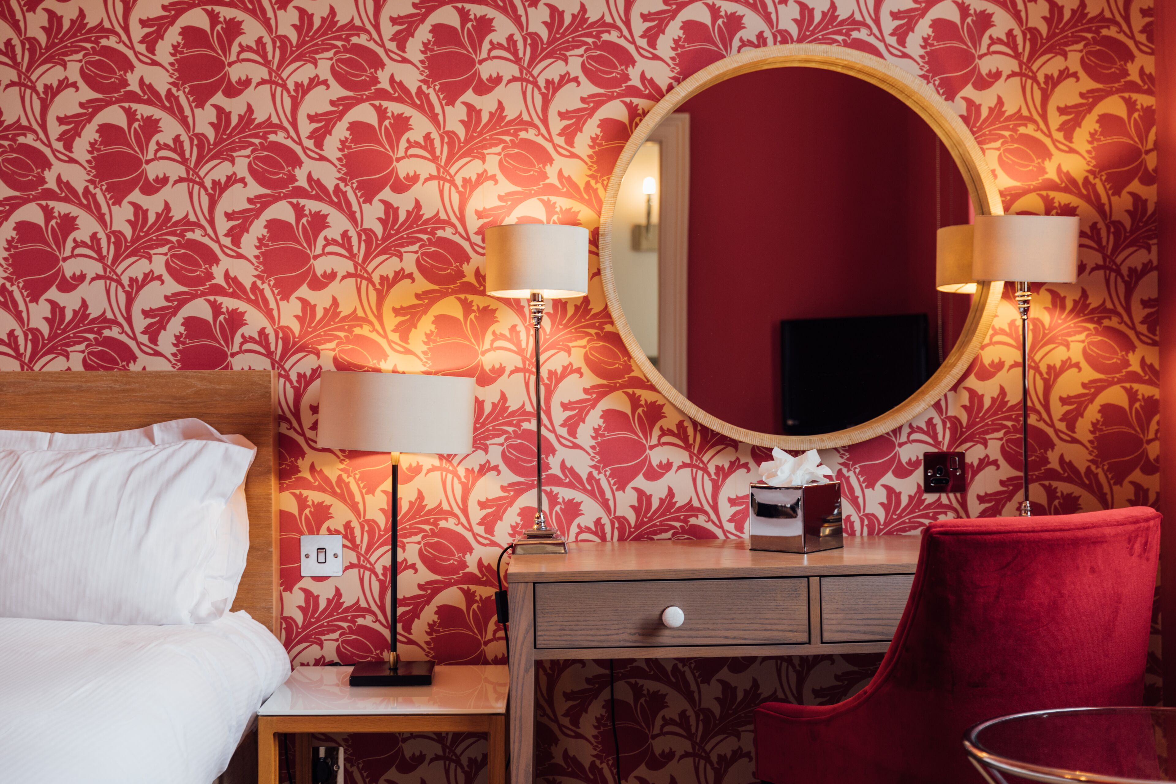 Quarto de hotel com cama, secretária com abajur, espelho e uma cadeira vermelha.