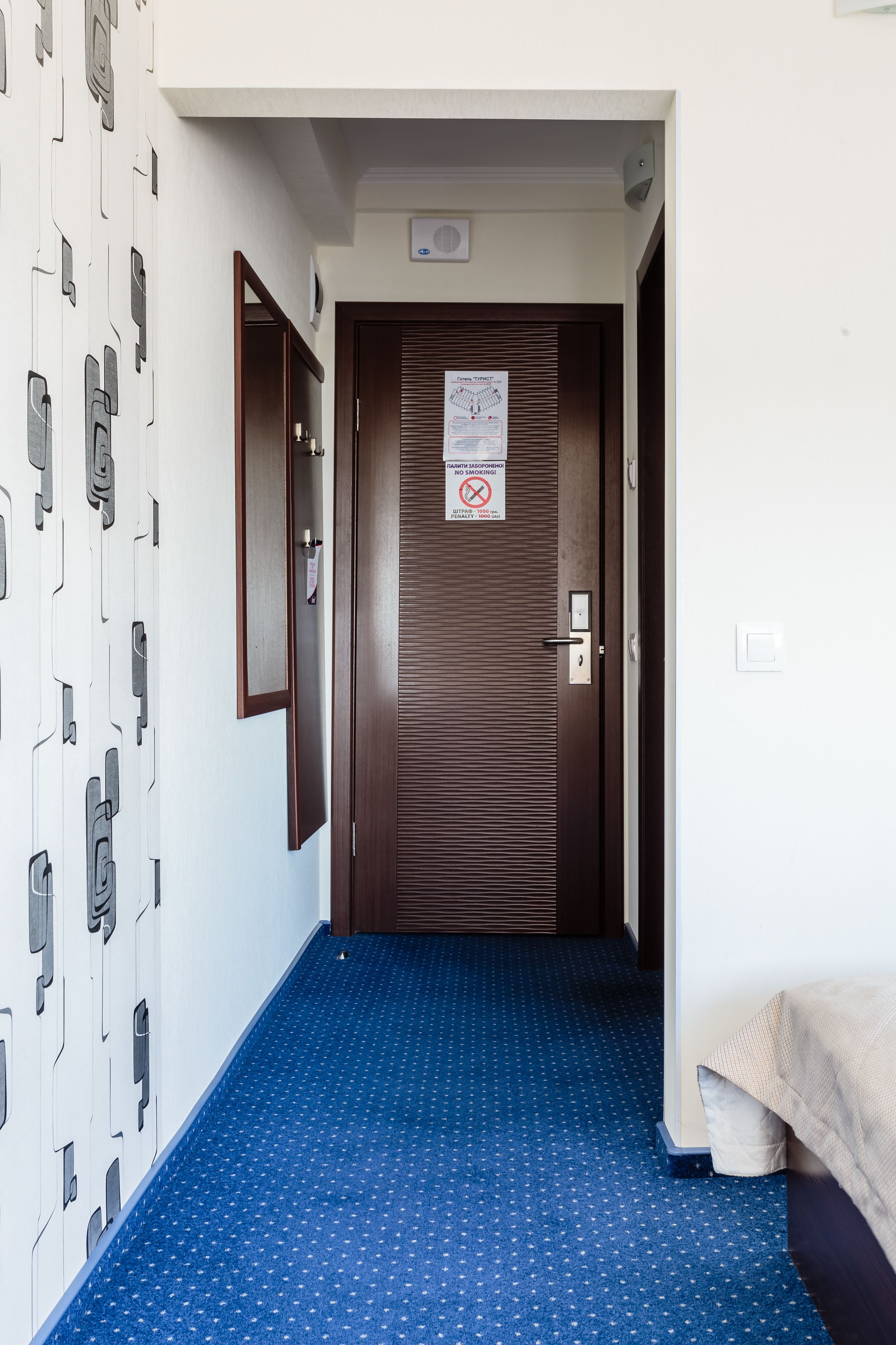 Corredor de hotel com carpete azul, porta de madeira com placa de segurança e parede com detalhes decorativos.