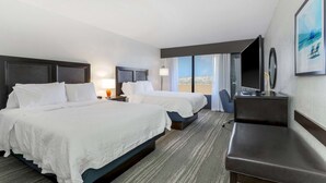 Quarto de hotel com duas camas, uma escrivaninha, televisão e uma janela grande com vista.