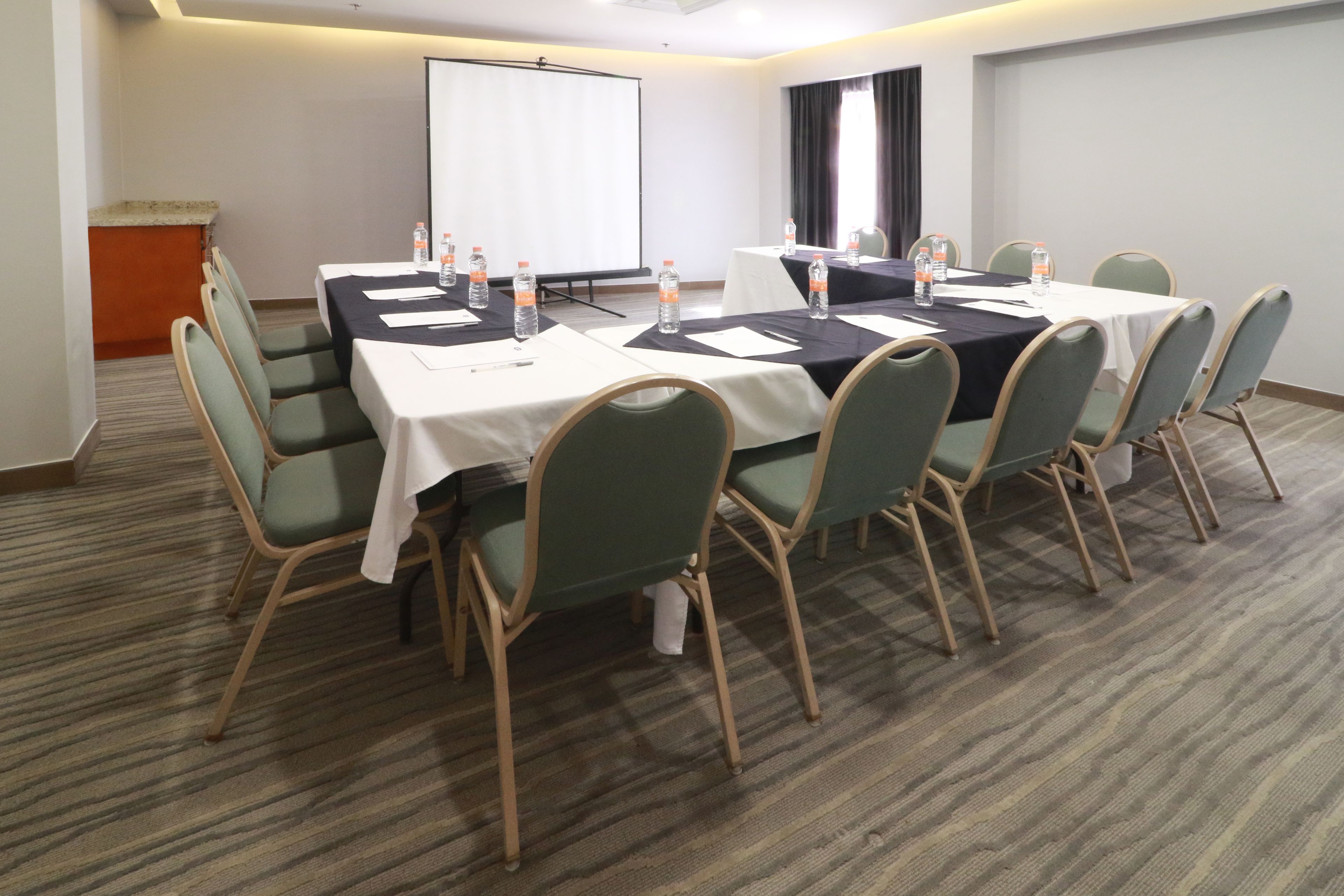 Sala de conferência com grande mesa, cadeiras, tela de projetor e garrafas de água.