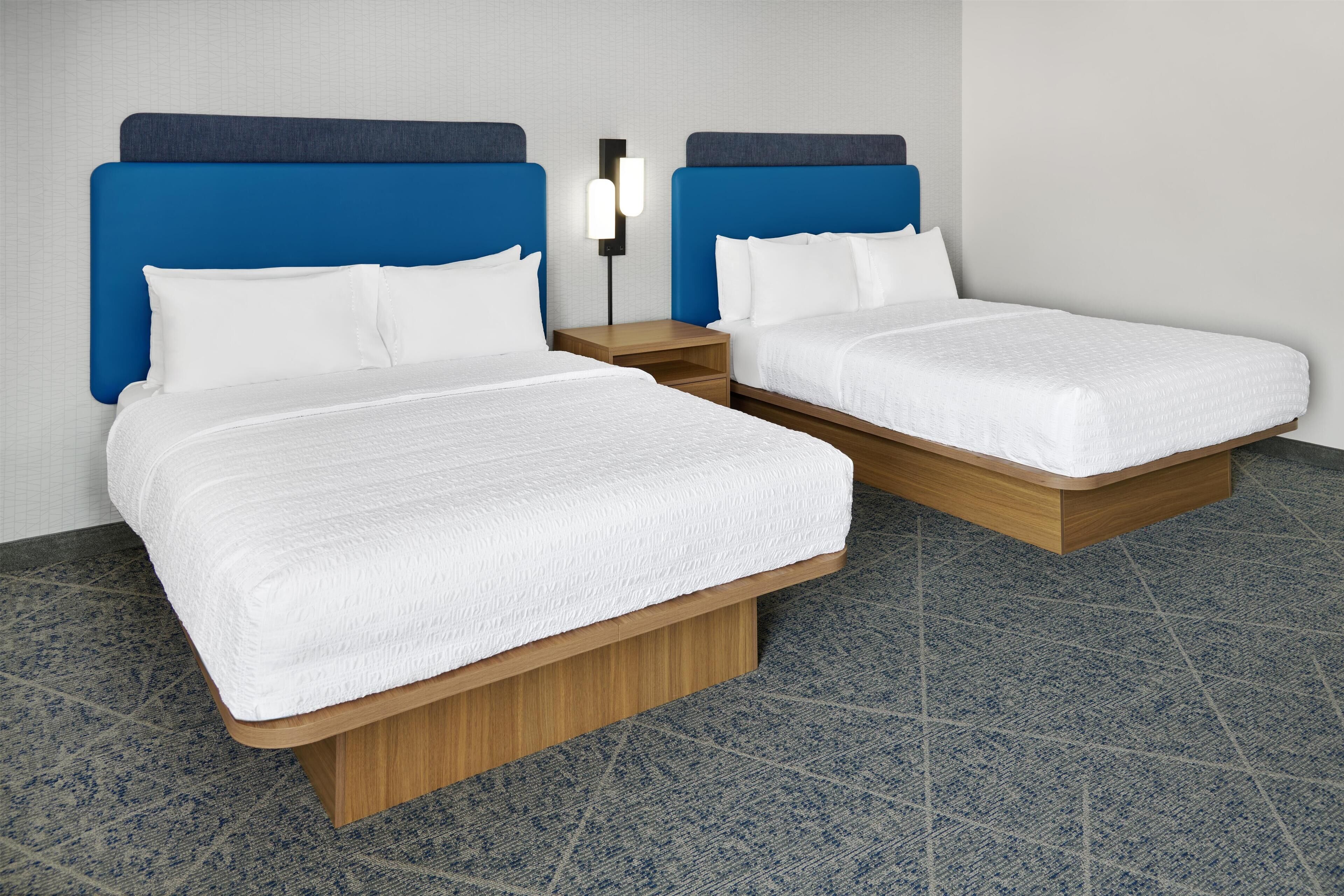 Quarto de hotel com duas camas de solteiro, cada uma com estrutura de madeira e cabeceiras azuis. Há uma mesa de cabeceira entre as camas com uma luminária sobre ela. O quarto possui um tapete azul com padrão geométrico.