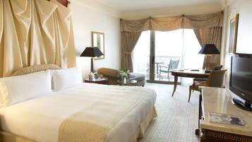 Quarto de hotel com uma cama grande, duas mesas de cabeceira, uma TV e varanda com mesa e cadeiras.