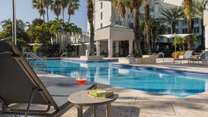 Área de piscina com espreguiçadeiras, uma mesa com bebida e um livro, e um edifício com telhado verde ao fundo.