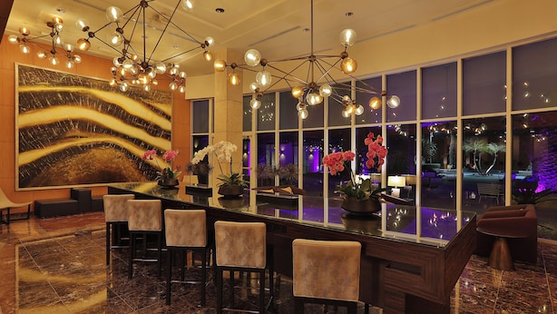 Um lobby de hotel moderno com piso de mármore, uma grande pintura abstrata, área de bar com banquetas e vista para a paisagem urbana através de amplas janelas.