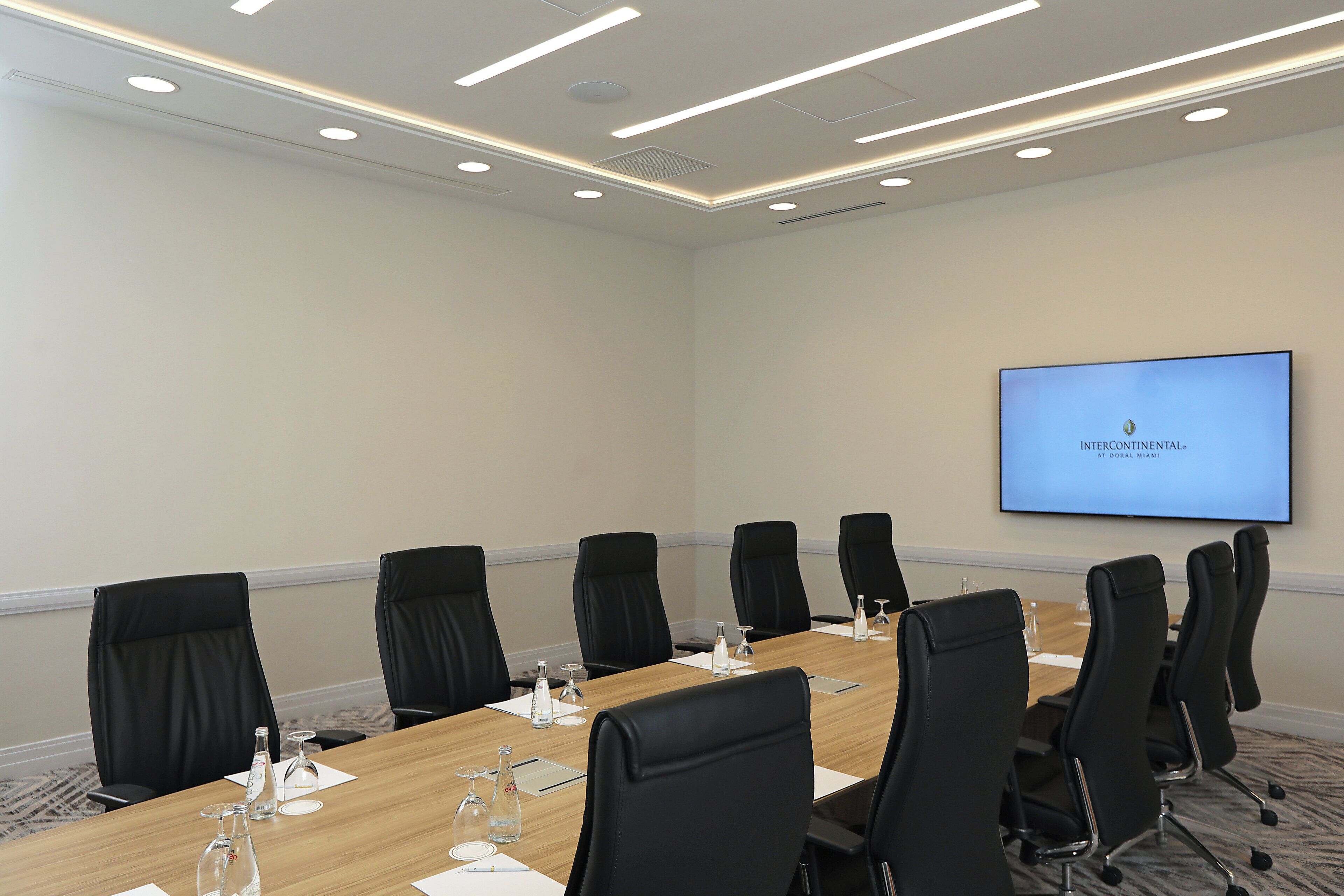 Uma sala de conferências com uma mesa comprida, cadeiras pretas e uma tela exibindo 'INTERCONTINENTAL AT DORAL MIAMI'.
