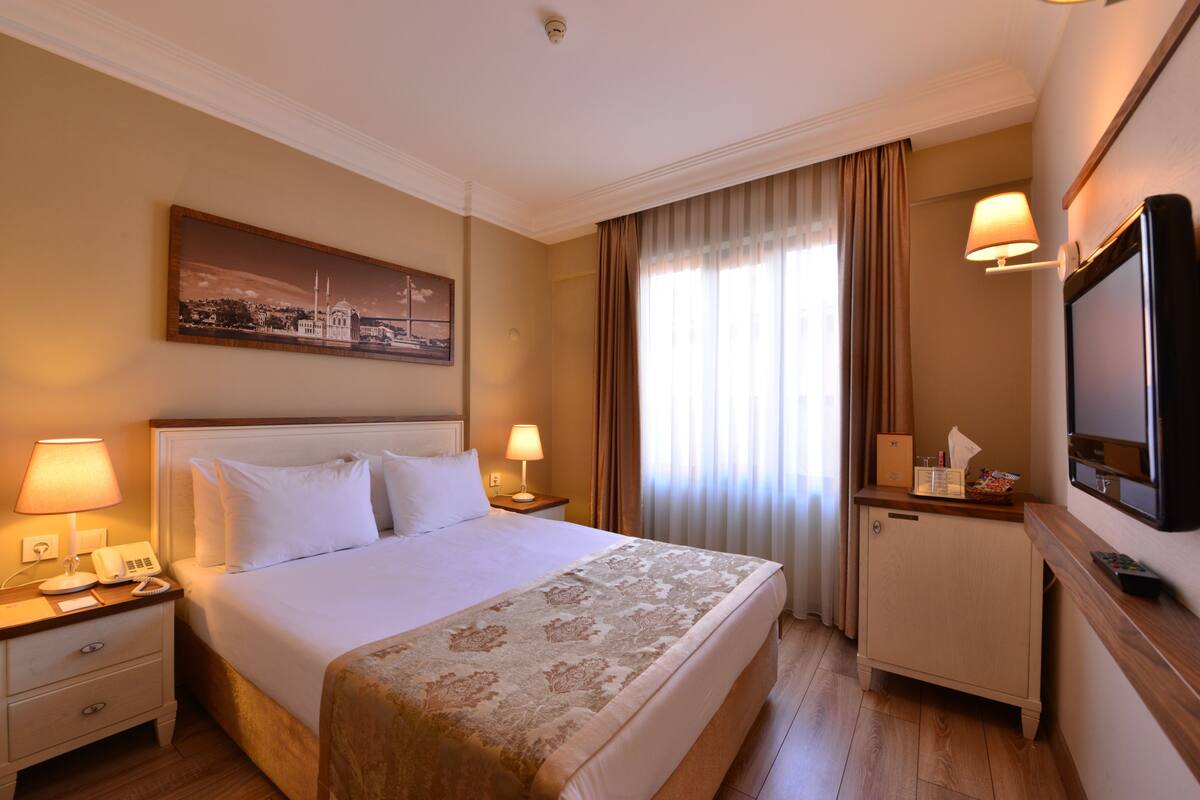 Quarto de hotel com uma cama grande, luminárias de cabeceira, uma pequena mesa de trabalho com luminária, televisão e um quadro na parede.