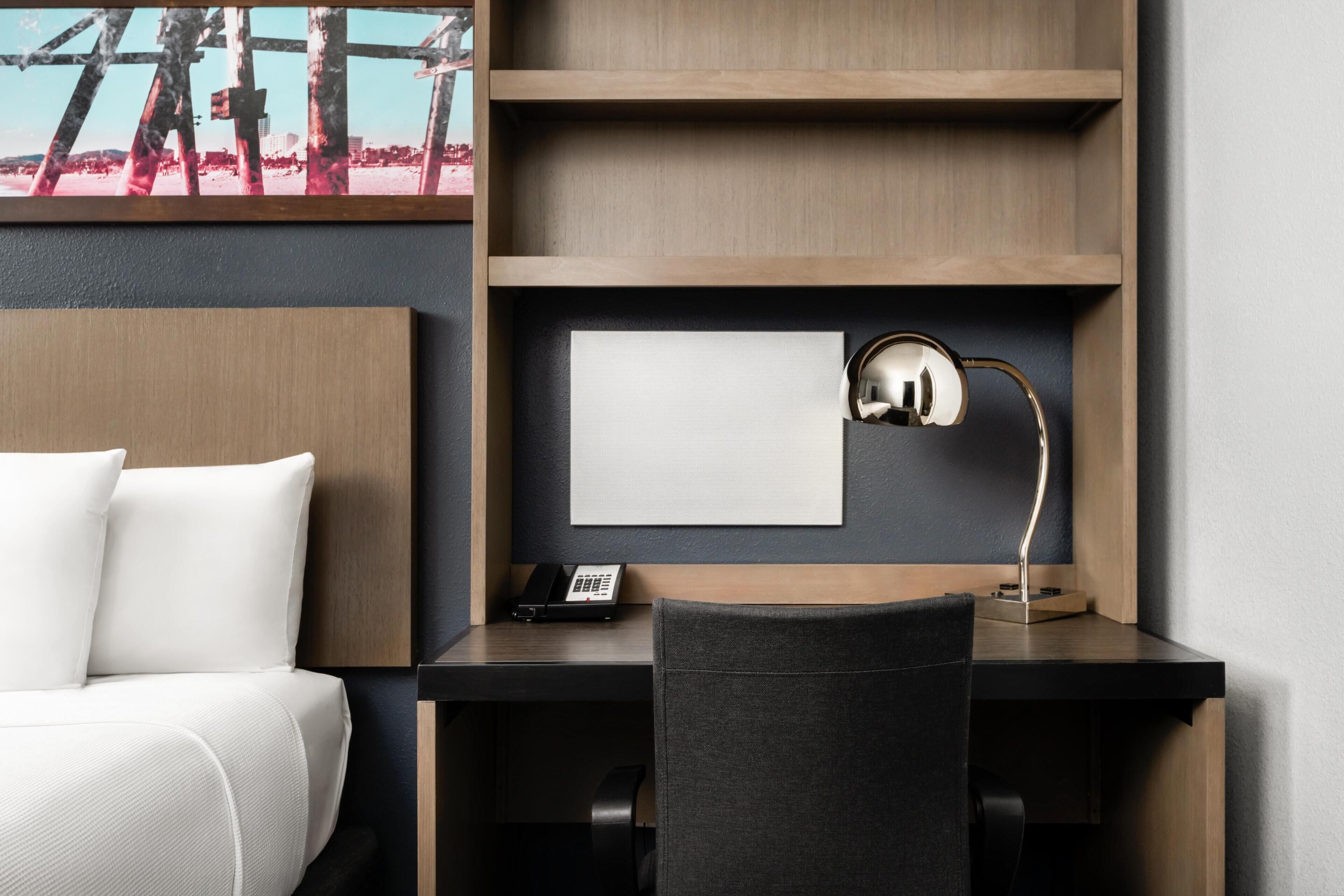 Quarto de hotel com cama, escrivaninha com luminária, telefone e quadro na parede.