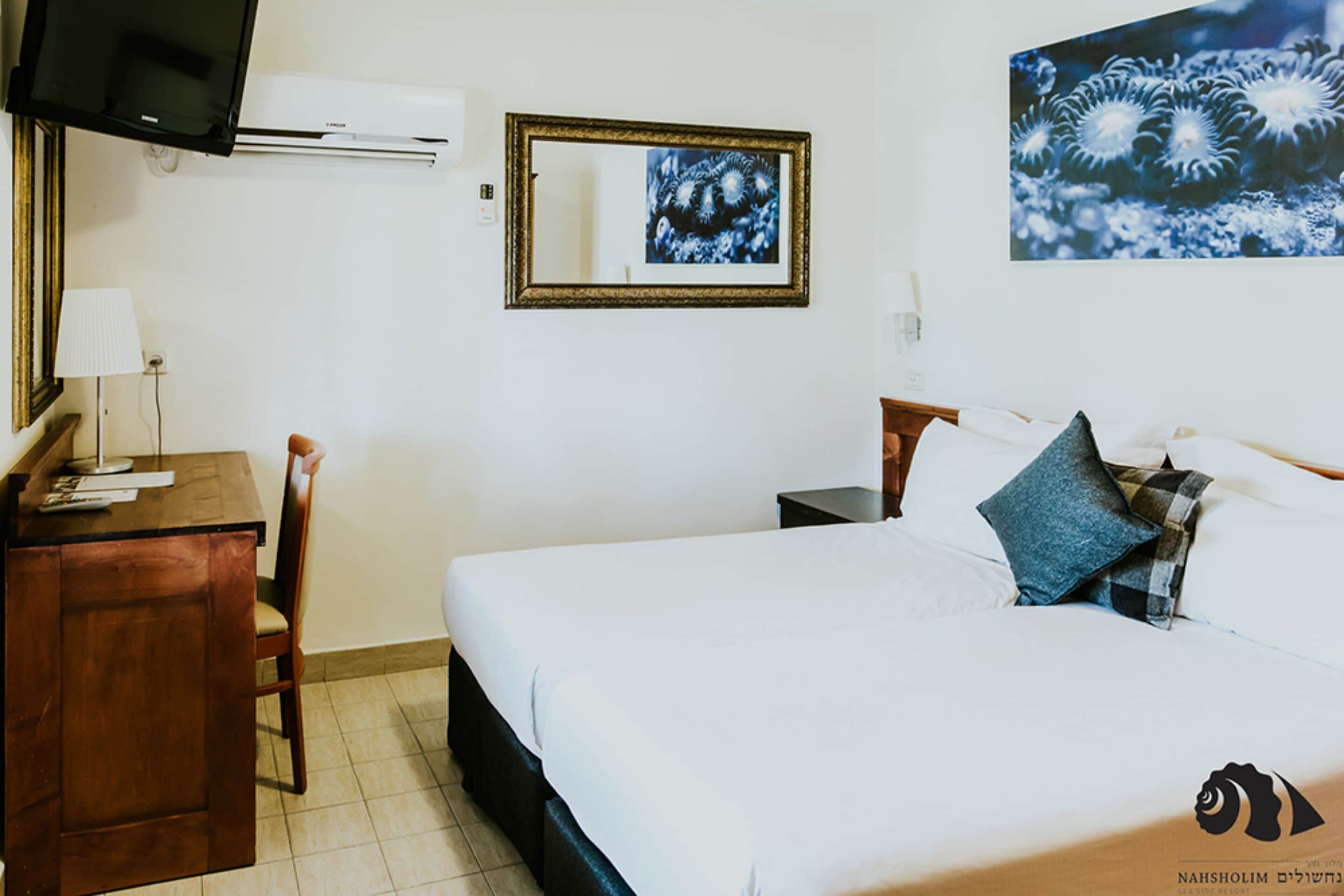 Quarto de hotel com uma cama, uma escrivaninha com cadeira, uma televisão fixada na parede e um quadro emoldurado sobre a cama.
