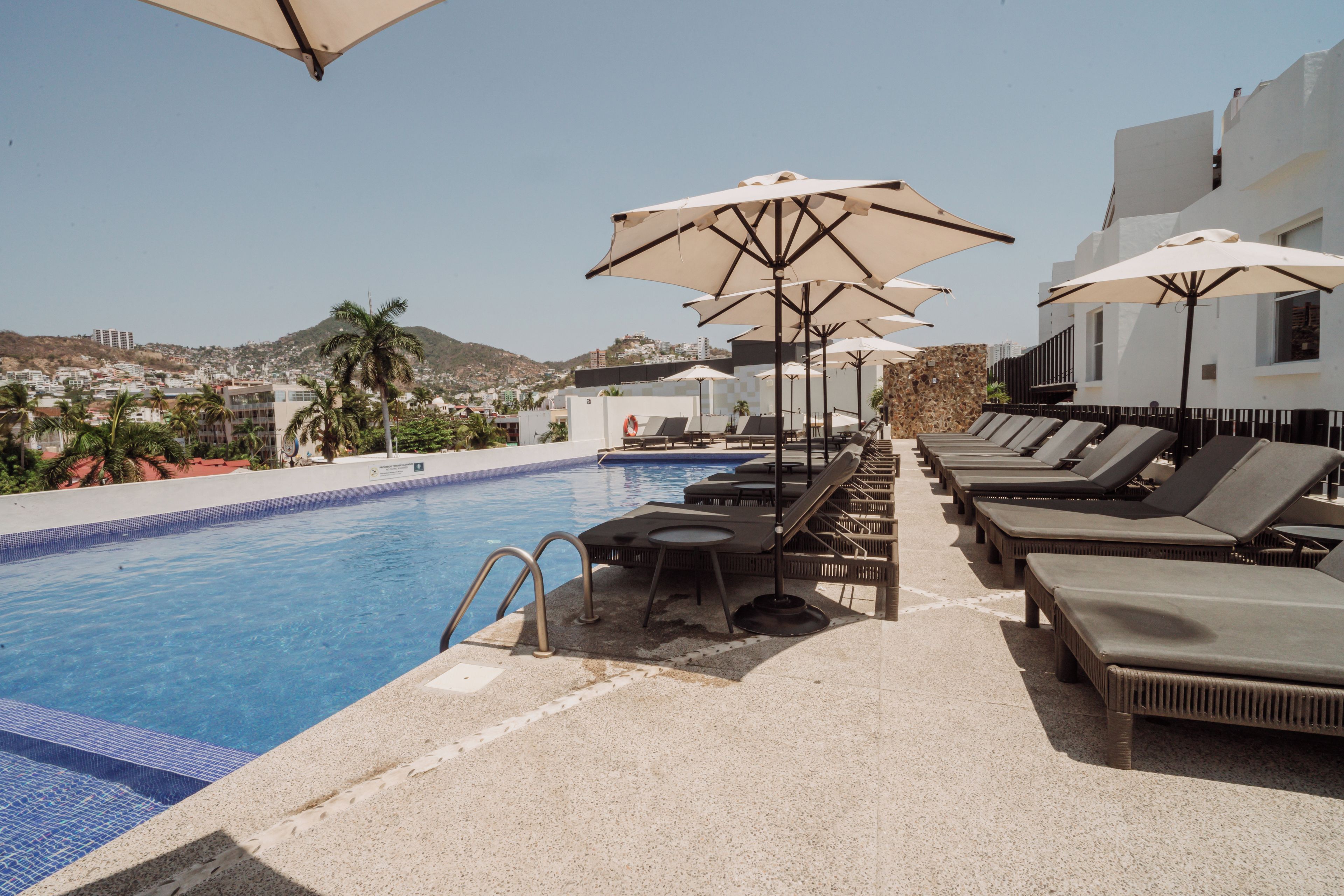 Área da piscina com espreguiçadeiras e guarda-sóis, piscina azul cristalina e vista para uma cidade no morro.