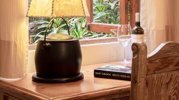 Uma mesa de madeira com uma luminária, um livro e uma garrafa de vinho perto de uma janela com vista para o verde.