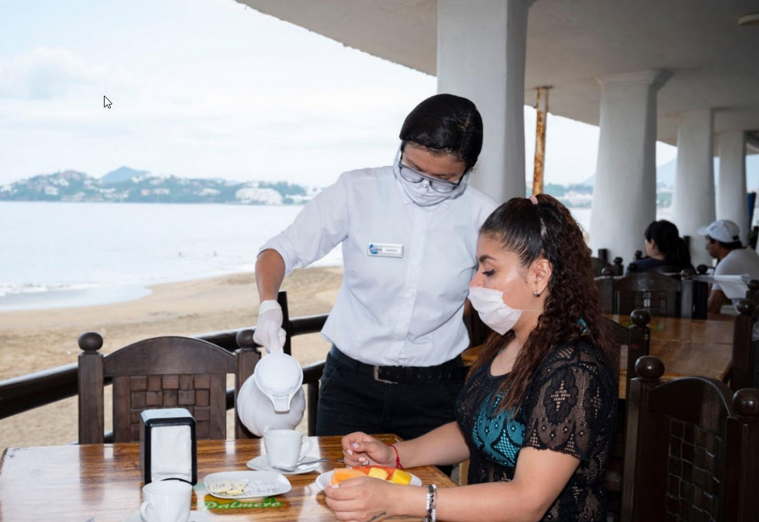 Uma área de refeições com uma mesa posta para dois, um garçom servindo uma refeição e vista para a praia ao fundo.