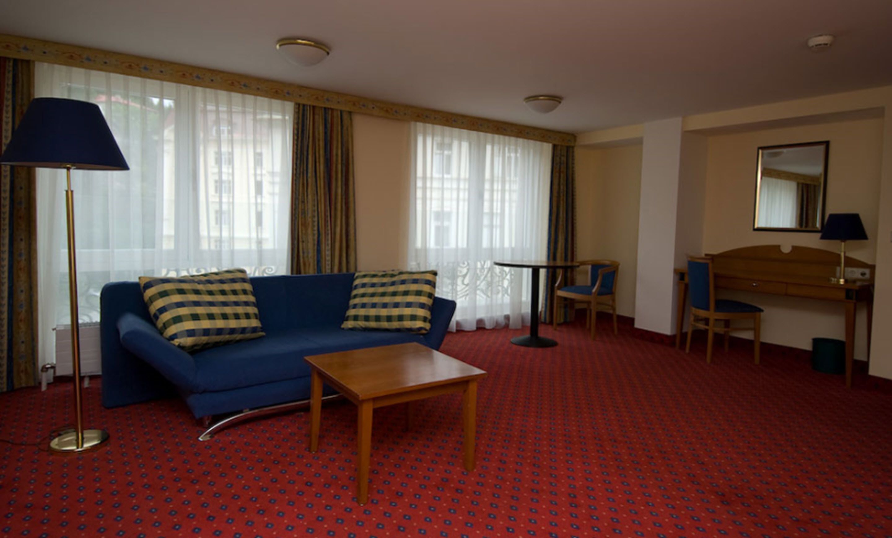 Quarto de hotel com um sofá azul, uma mesa de madeira, uma escrivaninha com cadeira e um espelho grande.