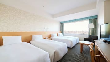 Quarto de hotel com três camas, uma escrivaninha, uma cadeira, uma televisão e uma janela com vista para a cidade.