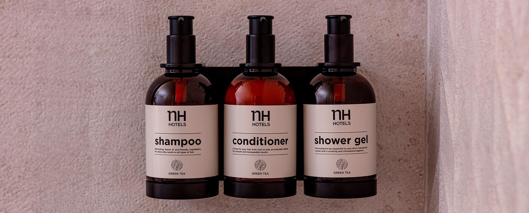 Três frascos de produtos NH Hotels: shampoo, condicionador e gel de banho, todos rotulados com 'GREEN TEA'.