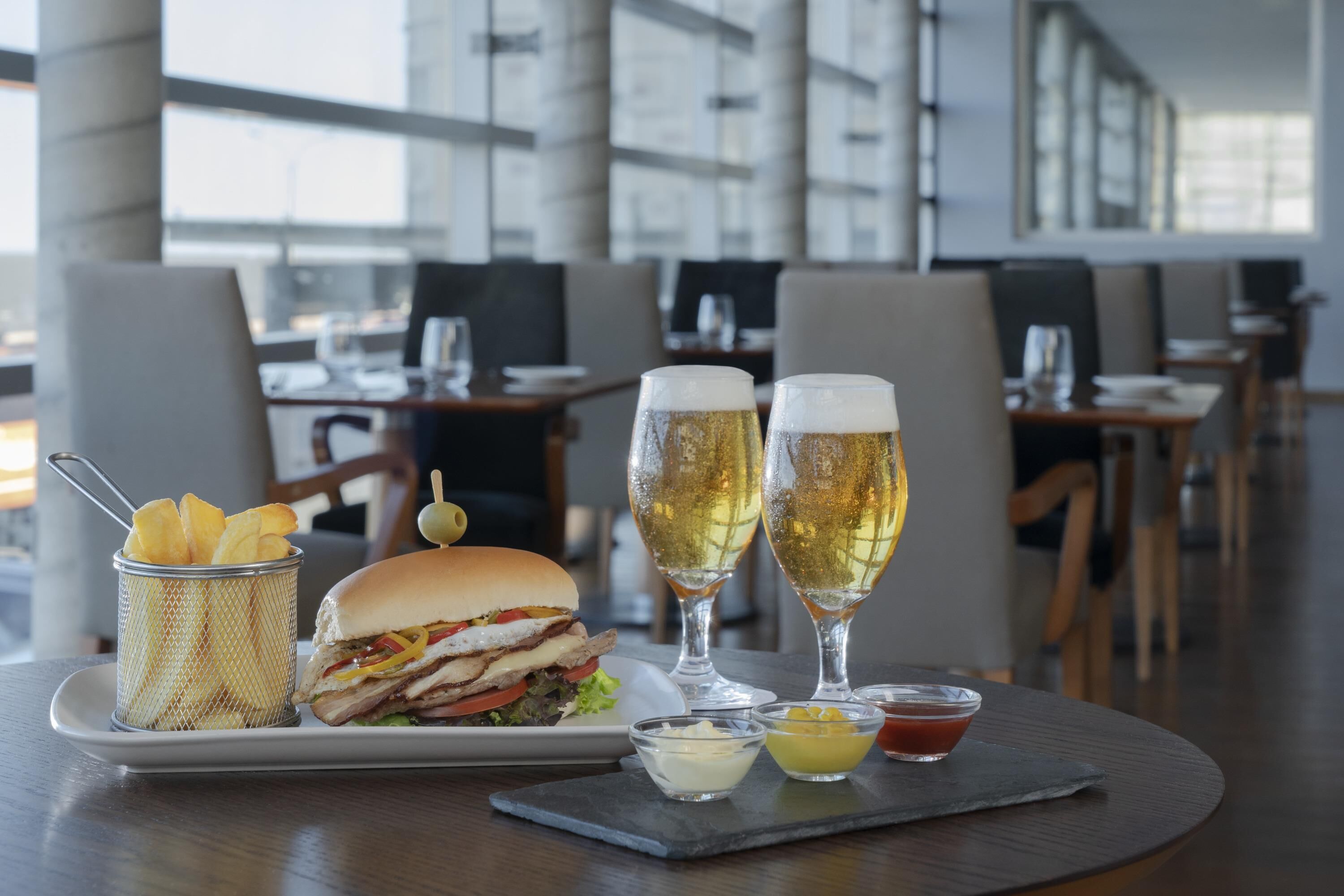 Uma mesa de jantar com um hambúrguer, duas cervejas, um cesto de batatas fritas e pequenos acompanhamentos.