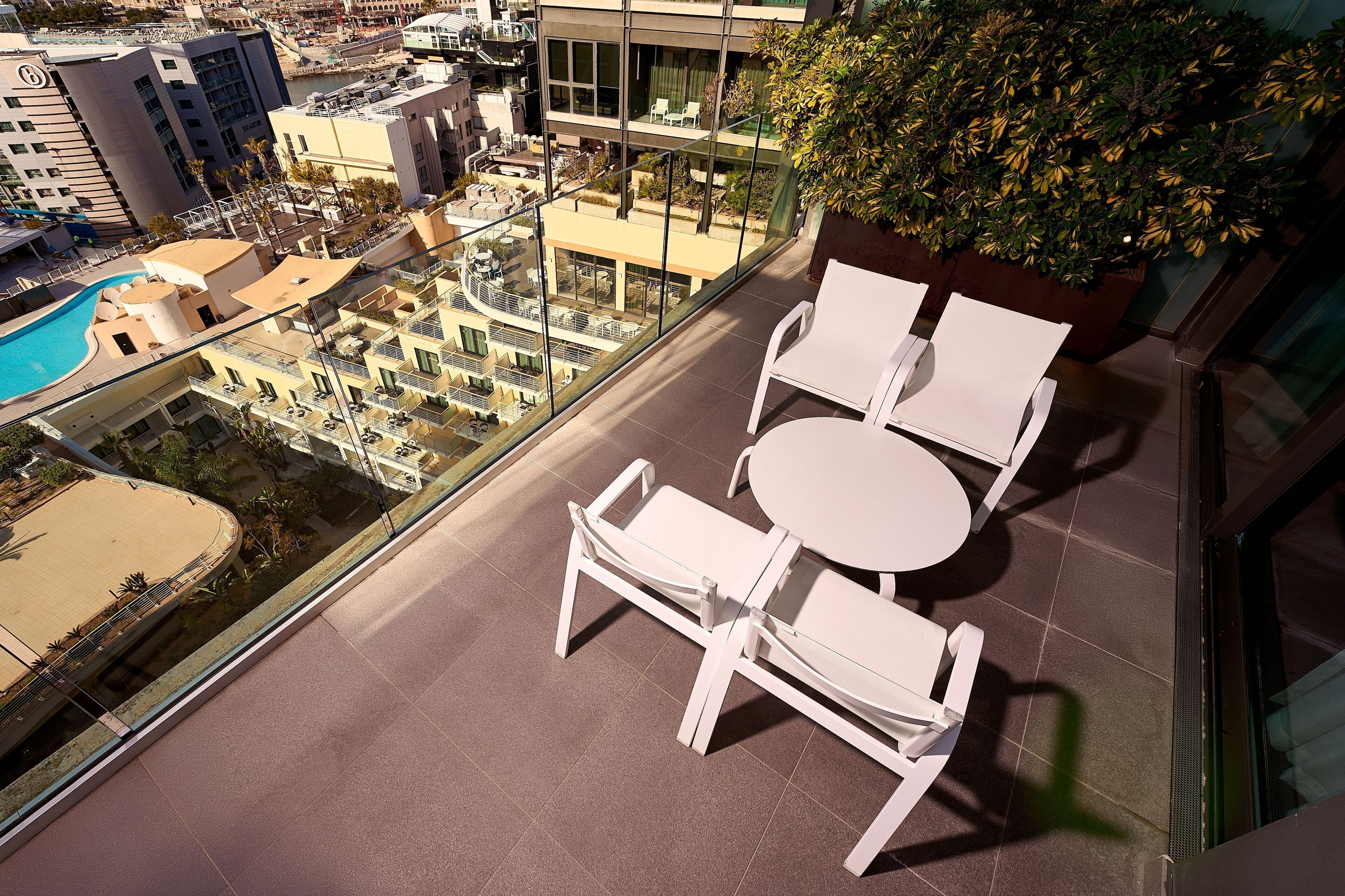 Varanda com cadeiras e mesa brancas, com vista para uma piscina e edifícios.
