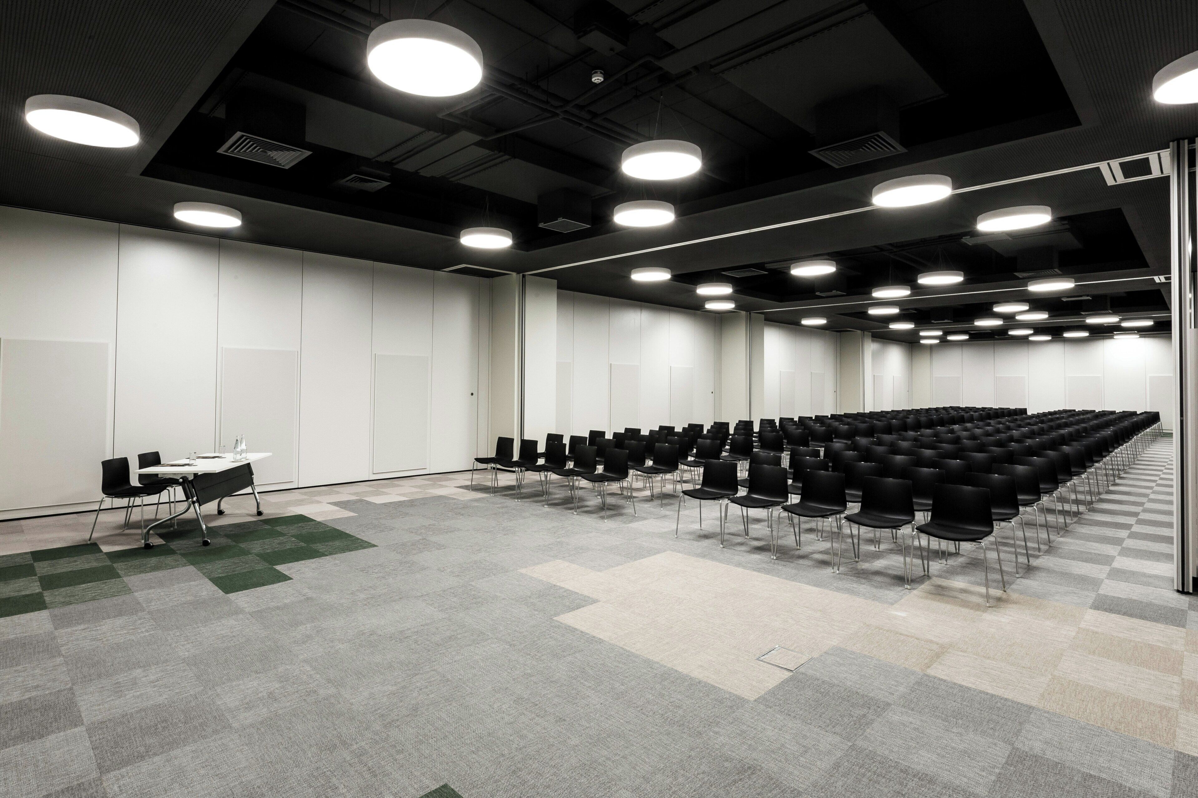 Uma sala de conferências com fileiras de cadeiras, uma mesa e cadeiras, e piso coberto por carpete.