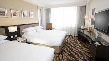 Quarto de hotel com duas camas, uma escrivaninha, uma televisão e uma janela grande com cortinas.