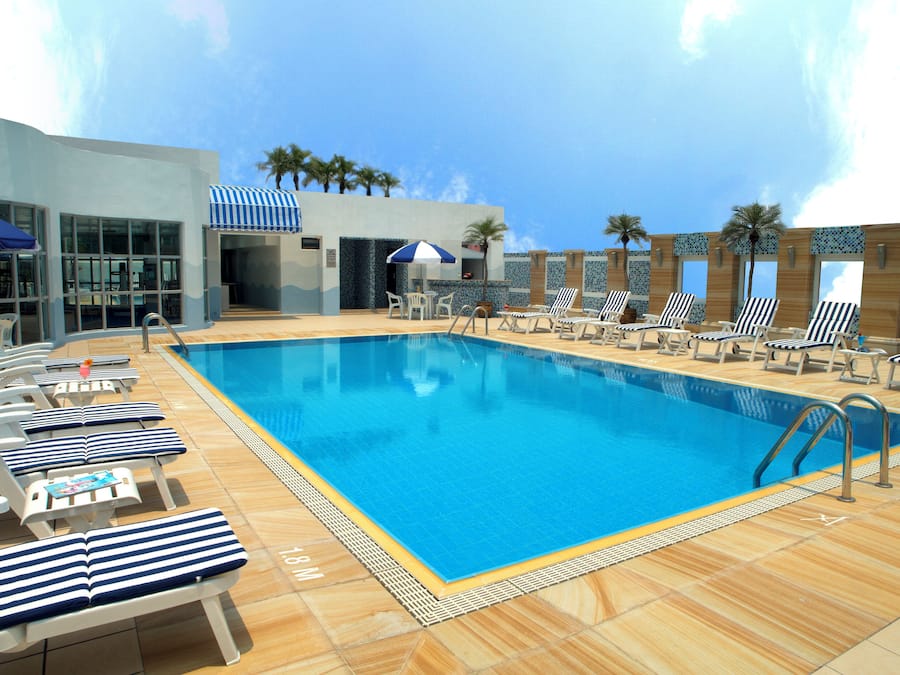 Piscina com espreguiçadeiras e guarda-sóis, cercada por um edifício com janelas amplas e palmeiras.