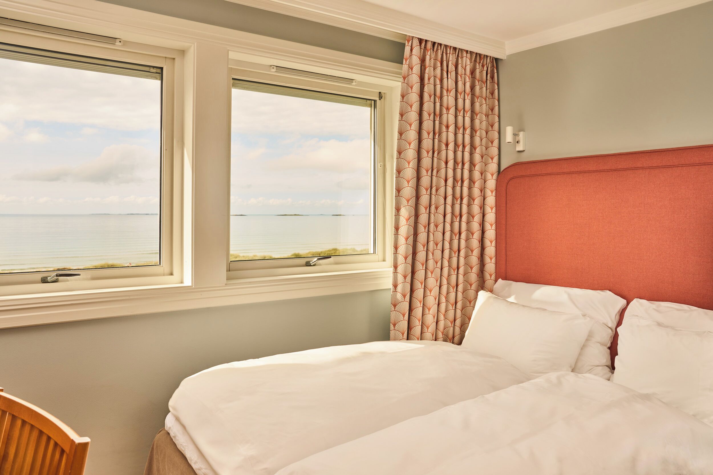 Quarto com cama, uma cadeira e duas janelas com vista para a água.