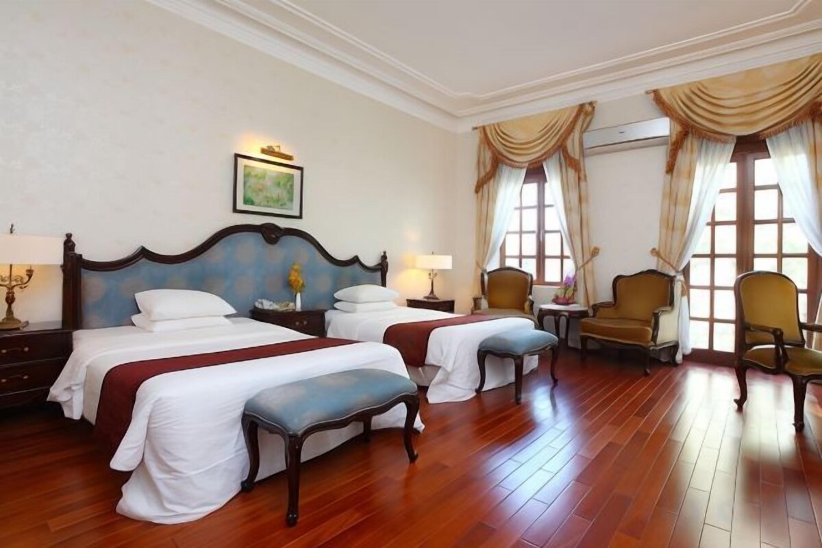 Quarto de hotel com duas camas, área de estar com cadeiras e uma janela grande com cortinas.