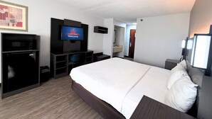 Quarto de hotel com cama, televisão na parede, pequena cozinha e banheiro visível através de uma porta aberta.