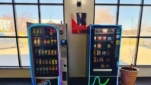 Duas máquinas automáticas com diversos snacks e bebidas, localizadas numa sala com grandes janelas.
