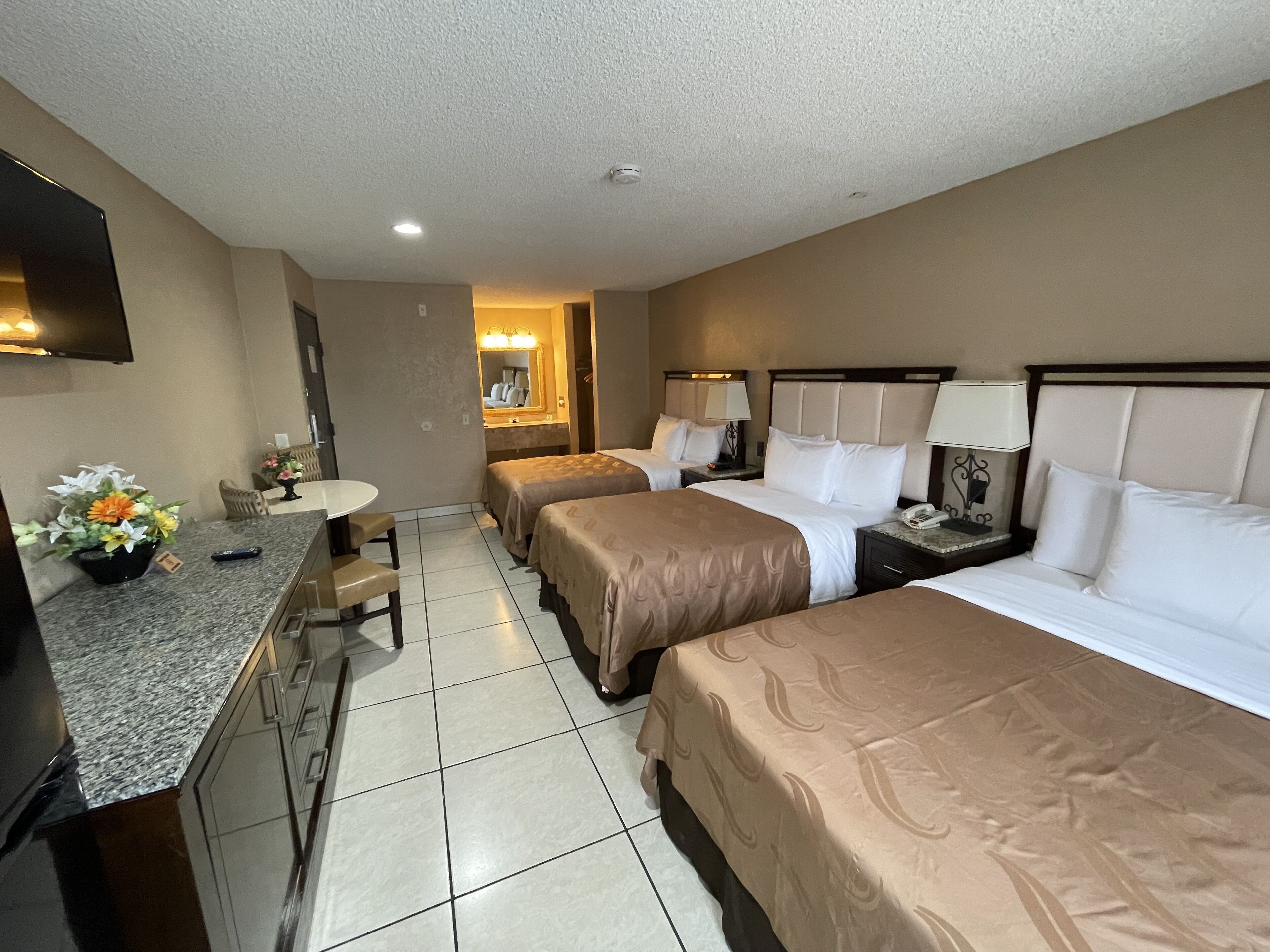 Quarto de hotel com duas camas, uma escrivaninha, uma cadeira, uma televisão e um banheiro visível por uma porta aberta.