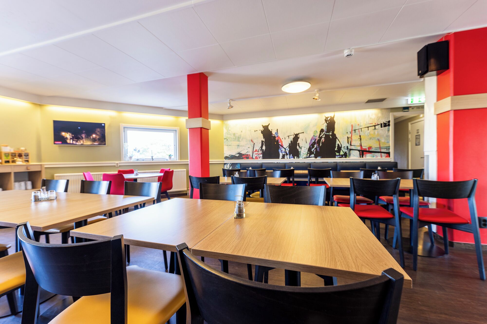 Um restaurante com várias mesas de madeira e cadeiras pretas, detalhes vermelhos nos pilares e paredes, e um grande quadro abstrato na parede do fundo.