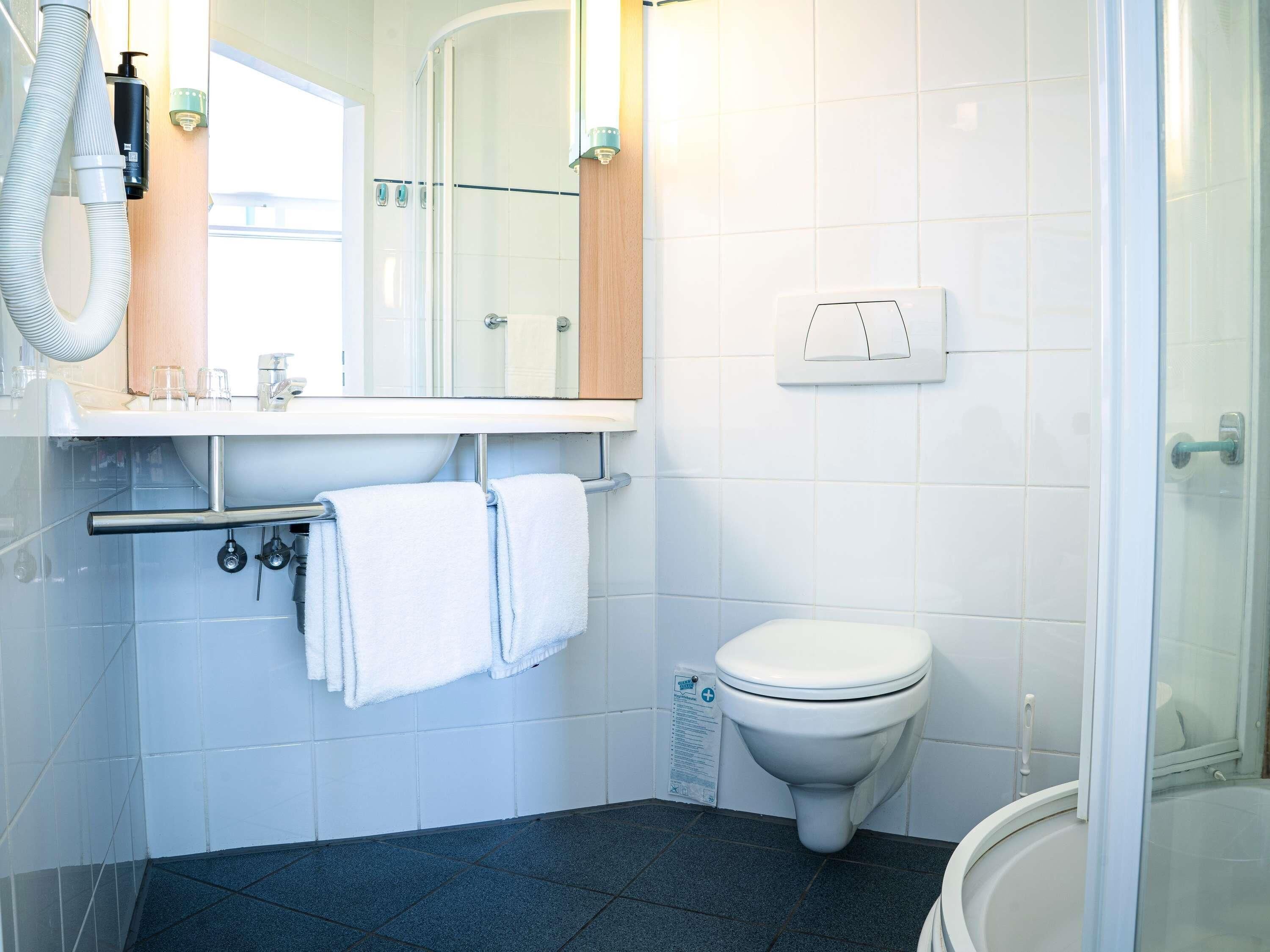 Um banheiro moderno com pia branca, espelho e vaso sanitário. Há um porta-toalhas com duas toalhas, uma área de chuveiro e um secador de mãos.