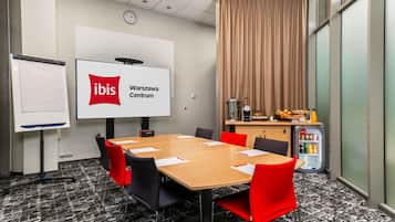 Uma sala de conferências com uma tela grande exibindo 'ibis Warszawa Centrum', uma mesa de madeira com cadeiras, um relógio, um quadro branco e uma geladeira com bebidas.