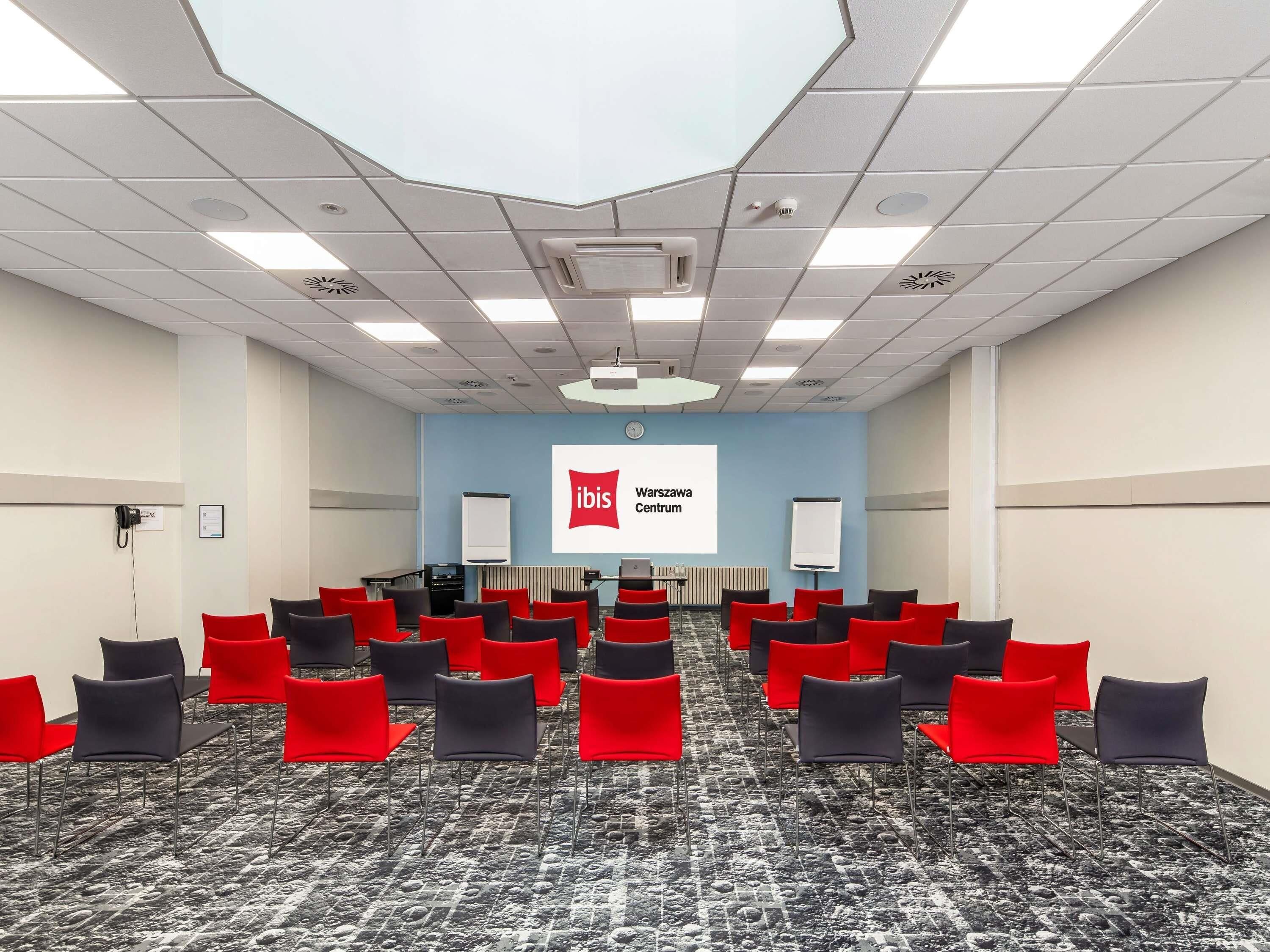 Uma sala de conferência com piso coberto por carpete, fileiras de cadeiras, uma tela de projetor e um quadro branco.
