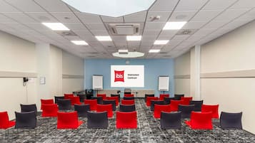 Uma sala de conferência com piso coberto por carpete, fileiras de cadeiras, uma tela de projetor e um quadro branco.