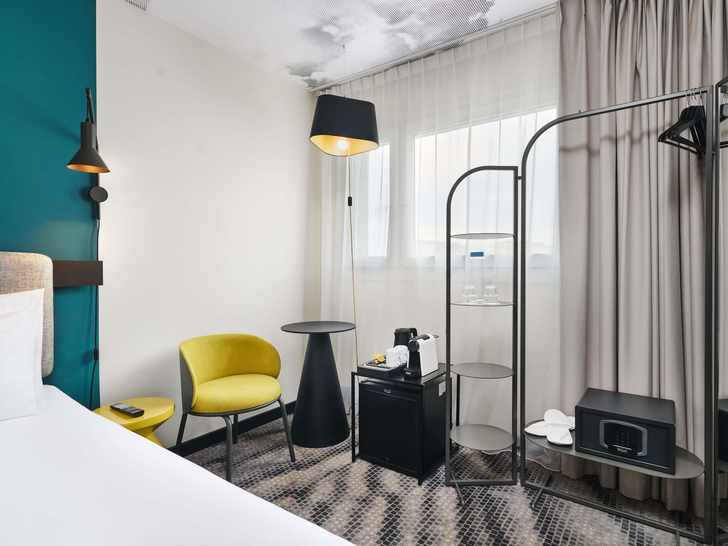 Quarto de hotel com cama, uma cadeira amarela, uma pequena mesa, abajur preto, armário preto, prateleira de vidro e alto-falante preto.