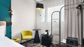 Quarto de hotel com cama, uma cadeira amarela, uma pequena mesa, abajur preto, armário preto, prateleira de vidro e alto-falante preto.