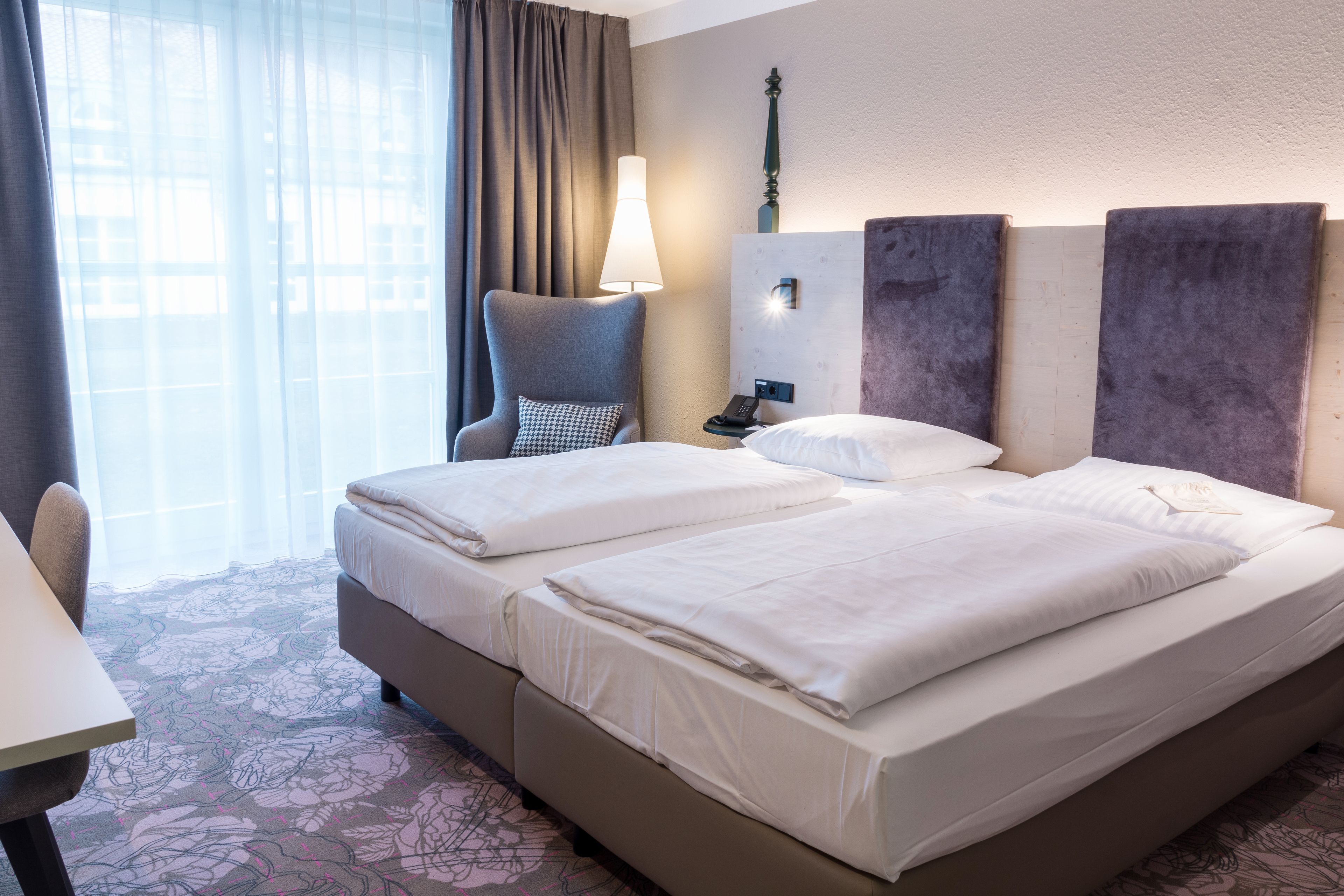 Quarto de hotel com uma cama grande, uma cadeira, uma escrivaninha e um abajur.
